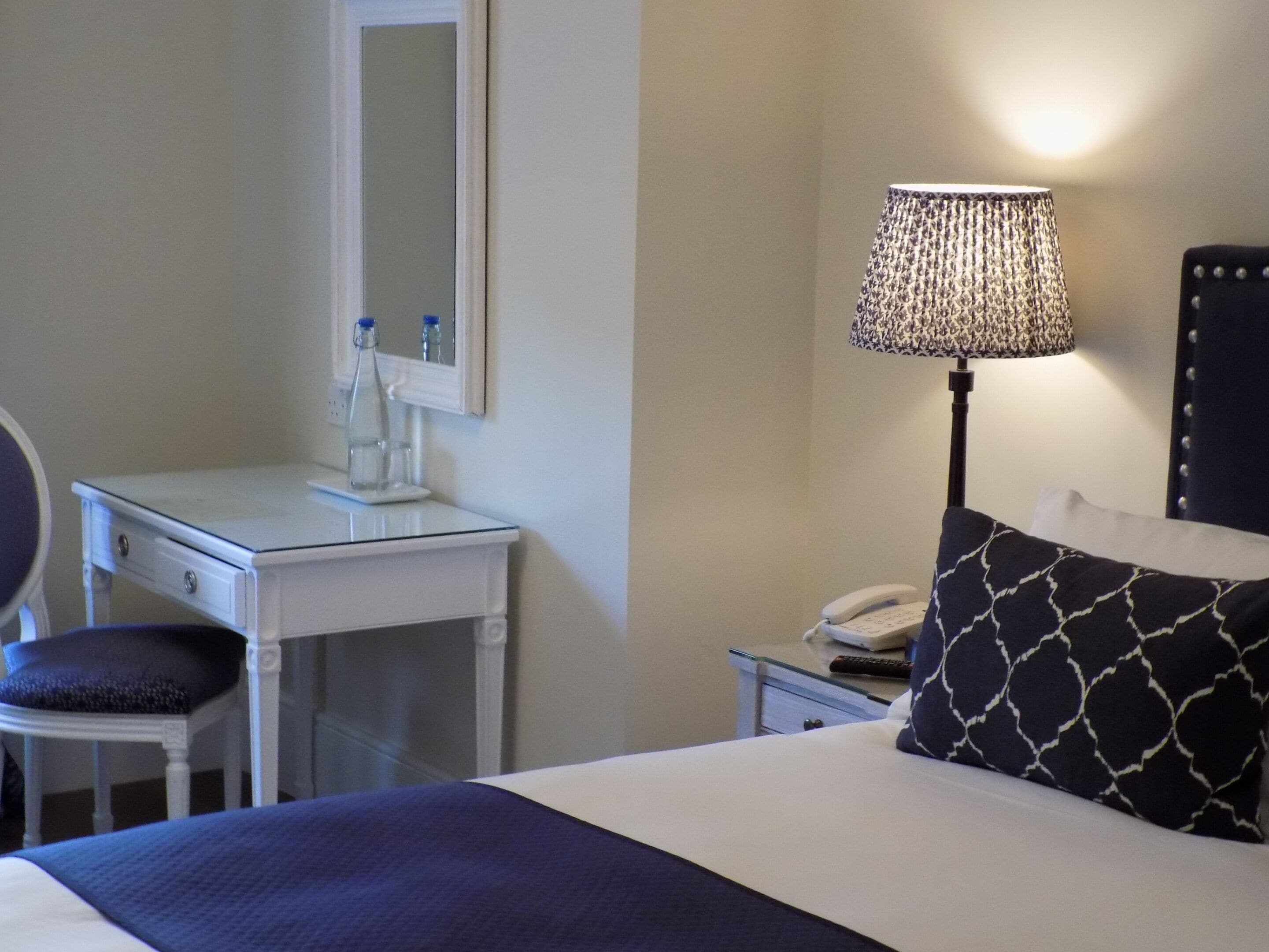 Quarto de hotel com cama, escrivaninha com espelho, luminária, cadeira e telefone em uma mesinha de cabeceira.