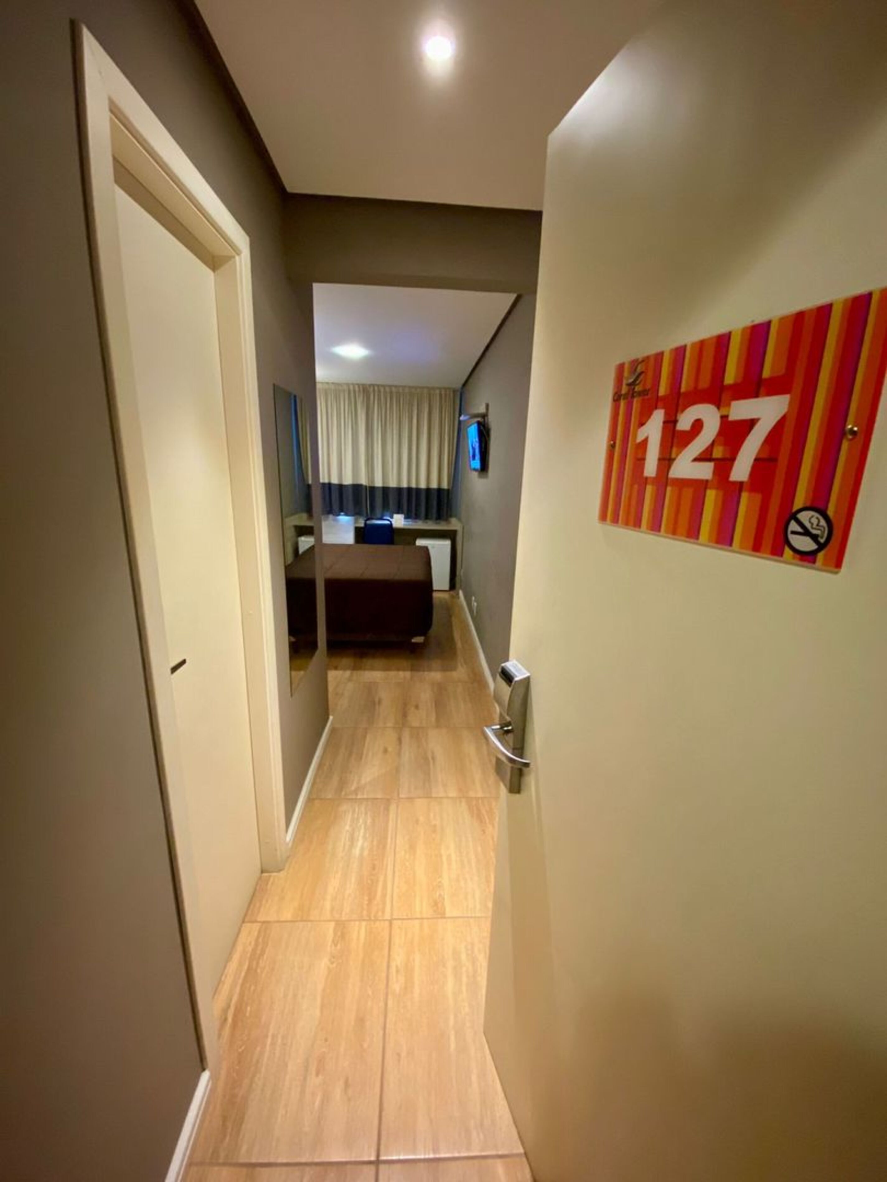 Corredor de hotel com uma cama, uma televisão e uma placa com o número do quarto.