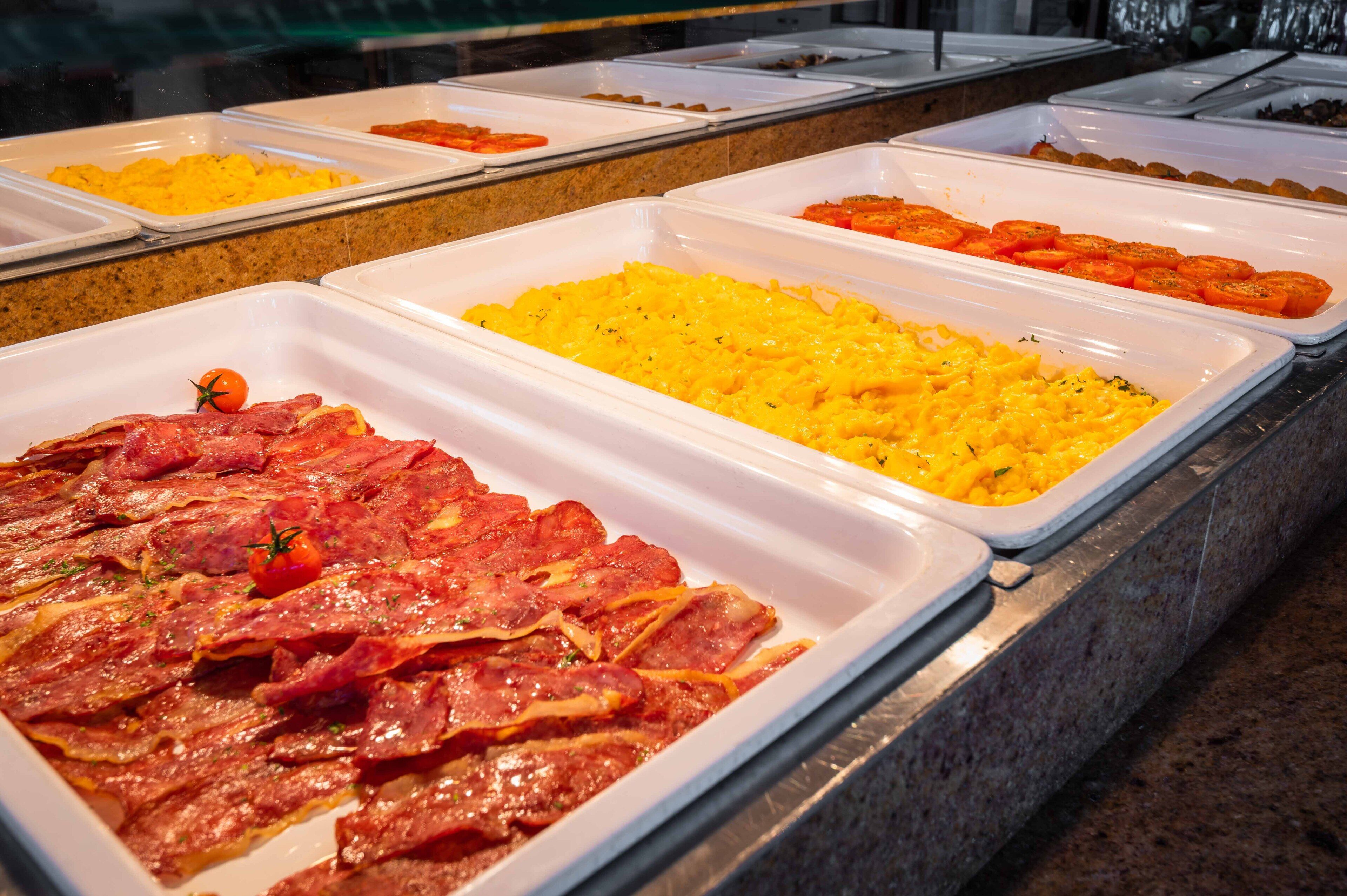 Buffet com diversas opções de comida, incluindo ovos mexidos, tomate e bacon.