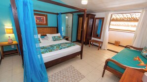 Uma cama de dossel com dossel azul, cabeceira de madeira e colcha verde-água. Há uma mesinha de cabeceira de madeira com abajur, uma cadeira de madeira e uma mesa pequena com um objeto decorativo.