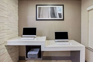Duas mesas com computadores, uma impressora e uma moldura com uma vista urbana na parede.