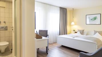 Quarto de hotel com cama, escrivaninha, cadeira, sanita e chuveiro.