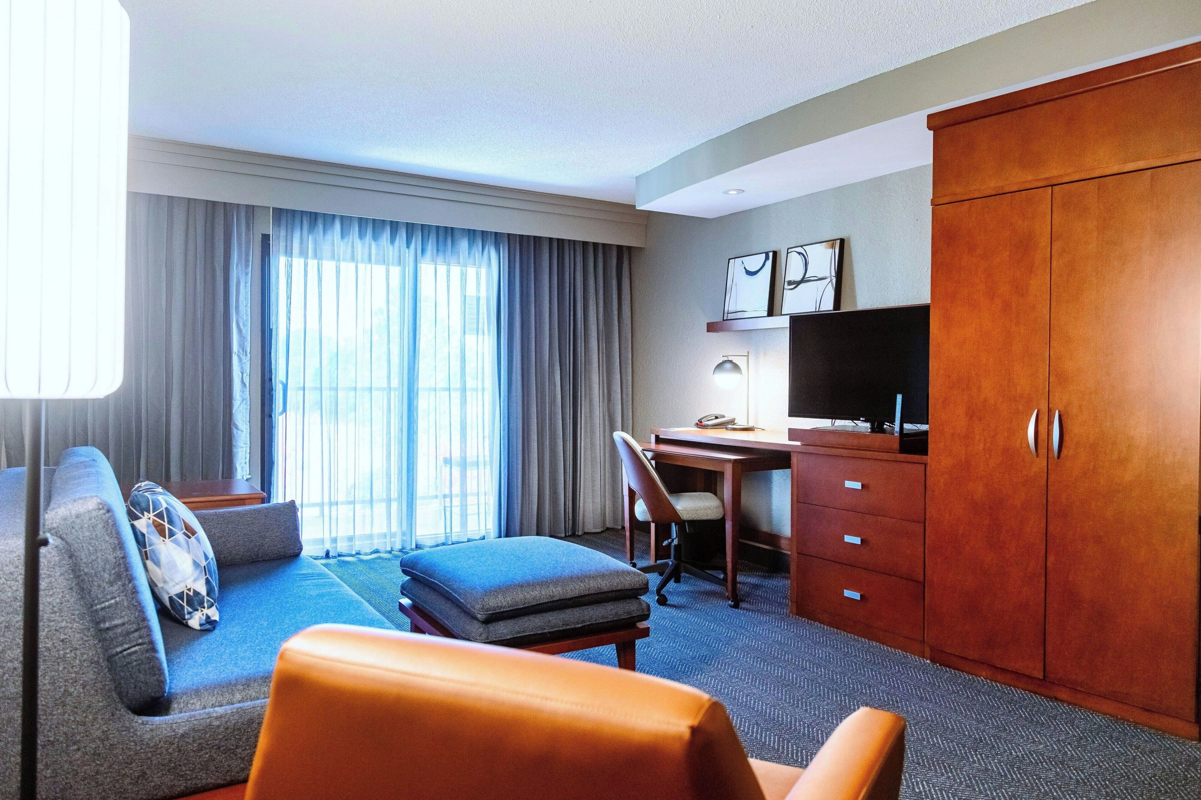Quarto de hotel com sofá, apoio para os pés, escrivaninha, televisão, guarda-roupa e cortinas.