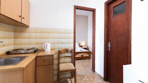 Cozinha compacta com pia, fogão e cadeira. Uma porta de madeira dá acesso a um quarto com cama e mesa de cabeceira.