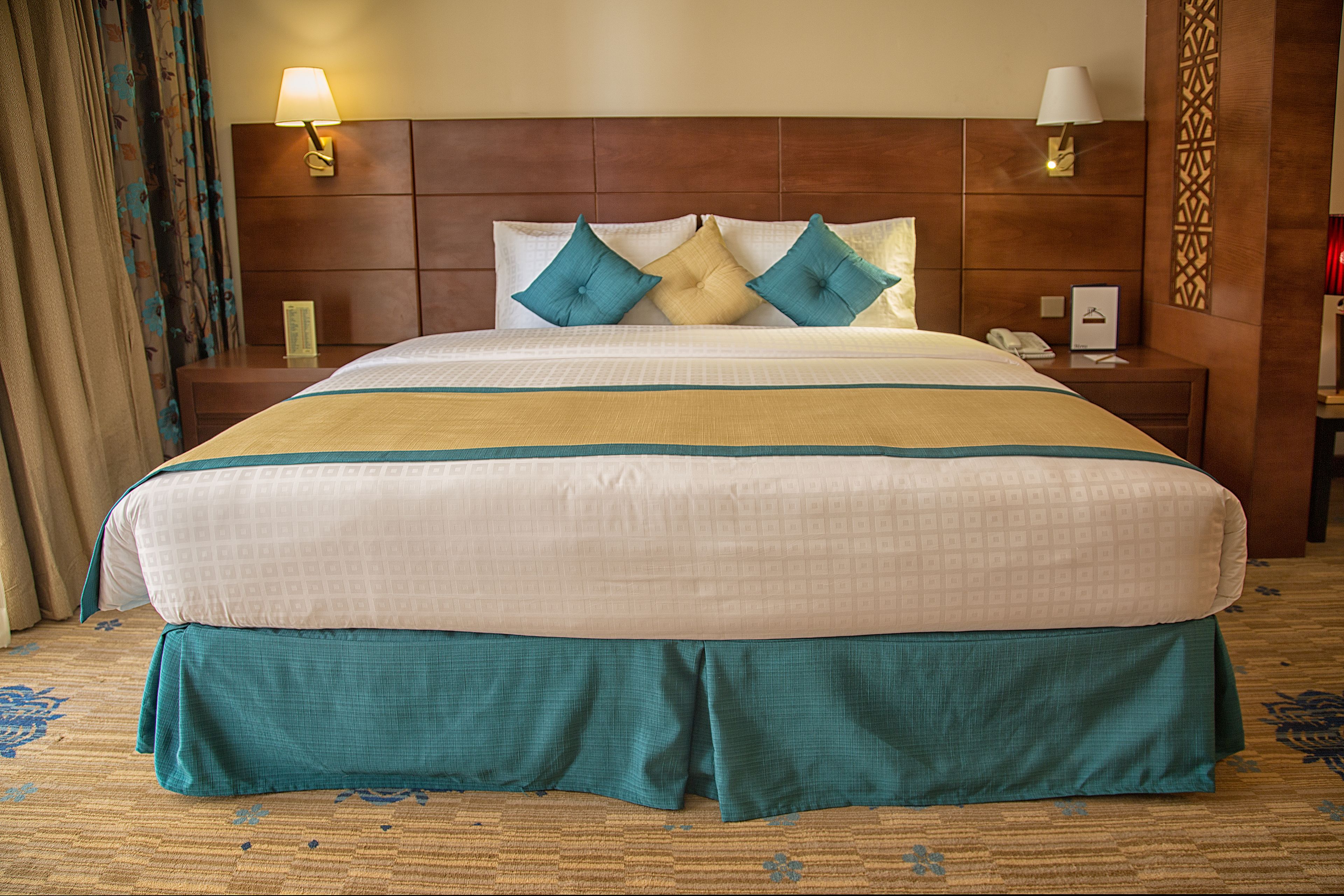 Quarto de hotel com uma cama grande, duas mesas de cabeceira, um criado-mudo e uma luminária na parede.