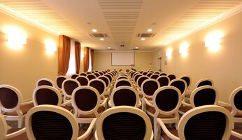 Uma sala de conferências com fileiras de cadeiras, uma tela de projetor e luminárias no teto.