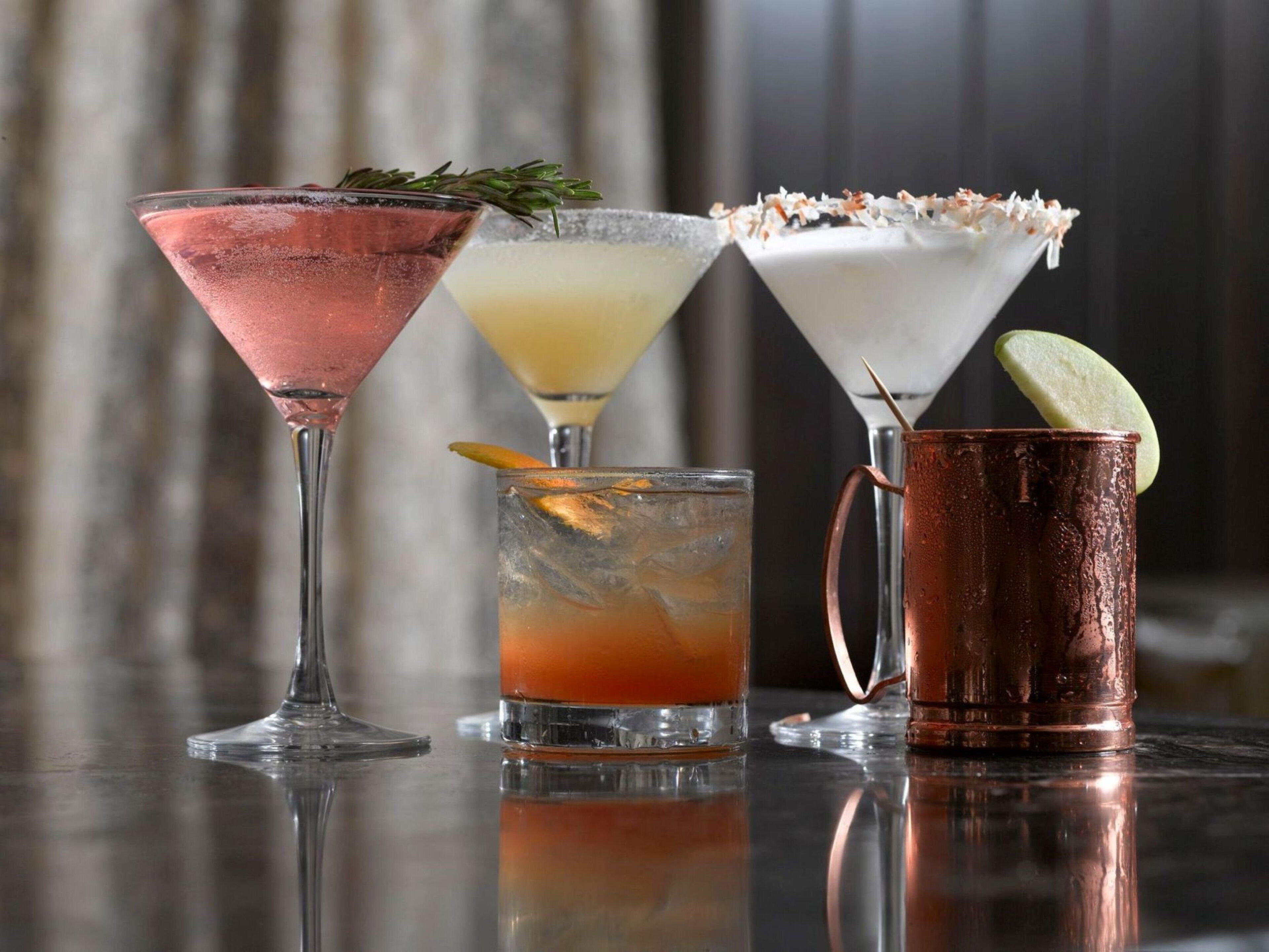 Quatro tipos diferentes de bebidas em diversos copos, incluindo um martini, um cocktail com guarnição de limão, uma margarita com borda salgada e um copo de cobre com uma fatia de limão.
