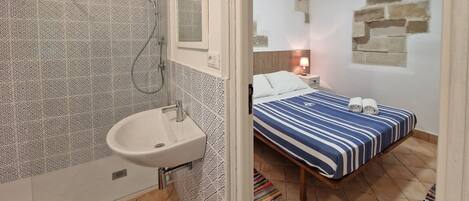 Casa de banho com lavatório, chuveiro e azulejos estampados. Uma cama com colcha listrada é visível no quarto adjacente.