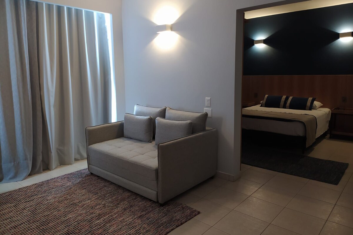 Quarto de hotel moderno com sofá cinzento, cama com cabeceira escura e luminária fixada na parede.