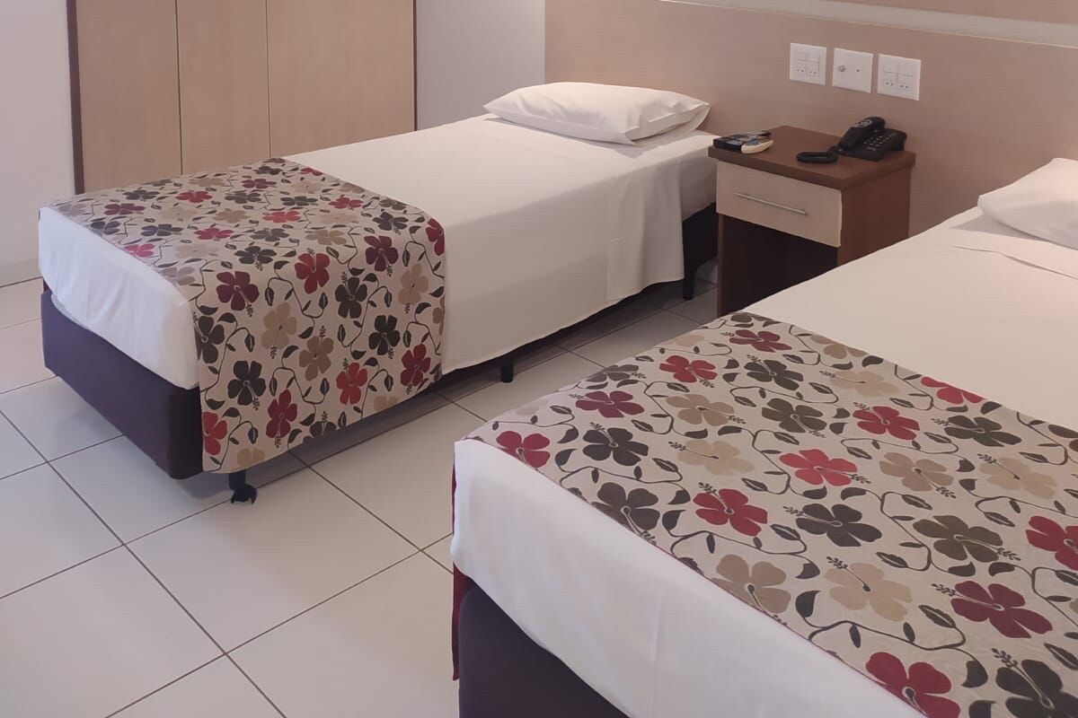 Quarto de hotel com duas camas de solteiro, um guarda-roupa, mesa de cabeceira com abajur e telefone.