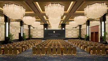 Uma sala de conferências com mesas e cadeiras dispostas em fileiras, grandes lustres e uma tela exibindo o nome do hotel.