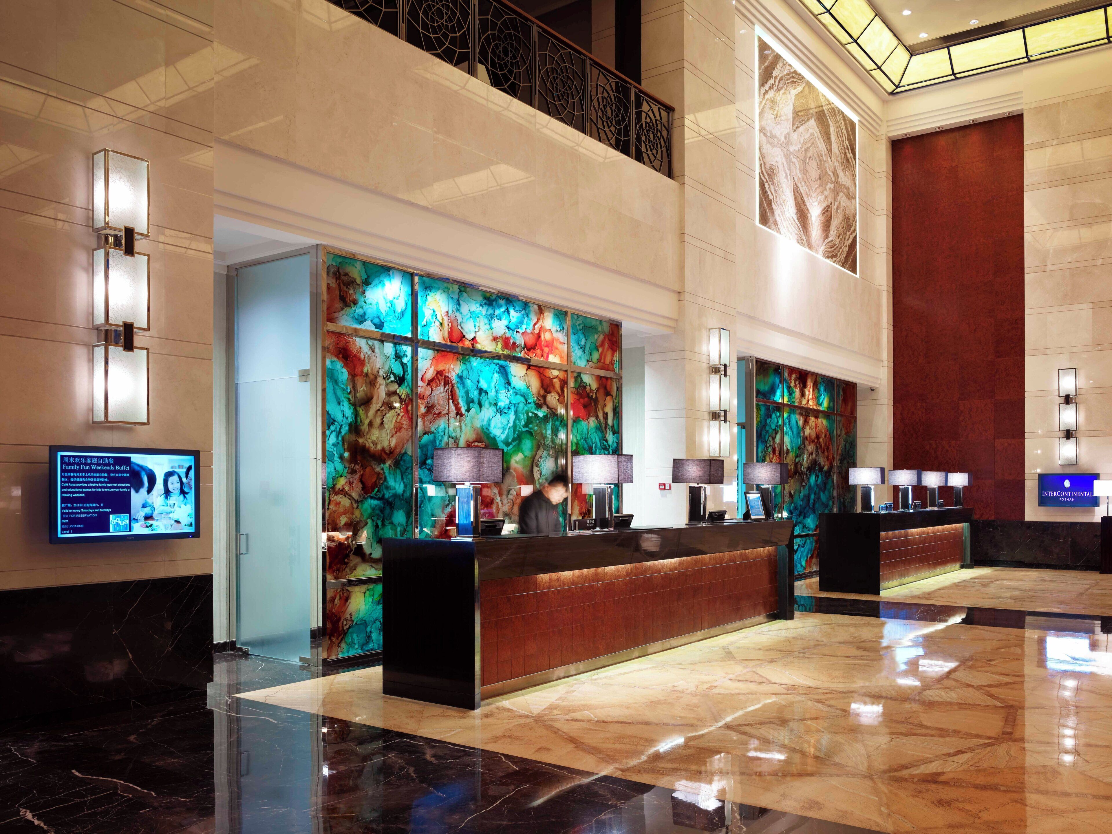 Uma área de recepção de hotel com uma grande obra de arte abstrata e colorida na parede, um balcão de atendimento com várias luminárias e um painel de informações digital.