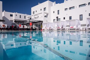Piscina com água azul cristalina, edifícios brancos e cadeiras vermelhas.