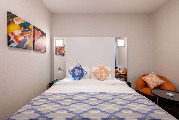 Quarto de hotel com uma cama, duas almofadas, uma cadeira, uma mesinha e duas pinturas na parede.