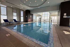 Piscina interna com espreguiçadeiras, grandes janelas e vista para a cidade.