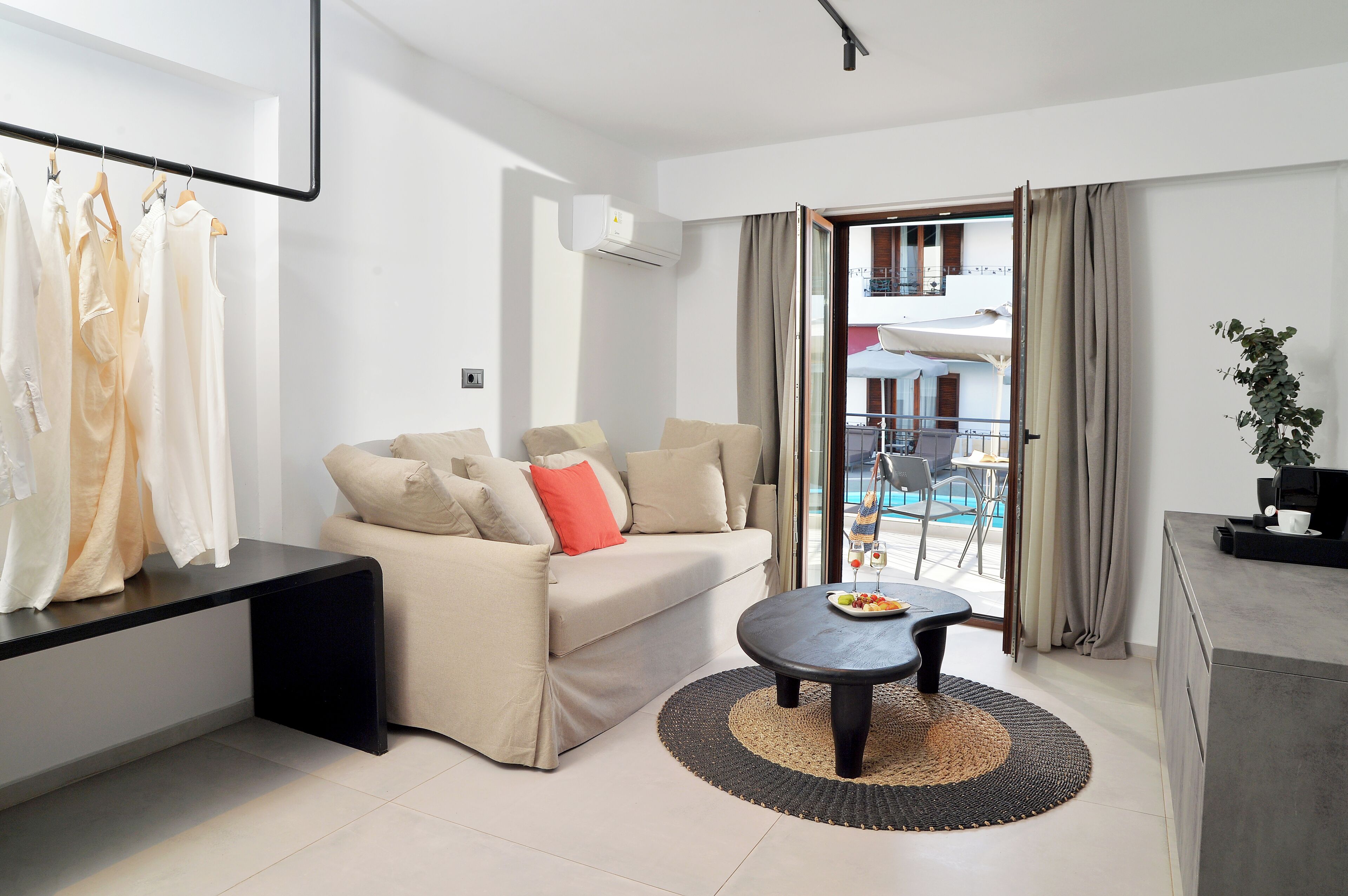 Sala de estar moderna com sofá, mesa de centro e um banquinho.  Varanda visível através das portas de vidro, com uma piscina.