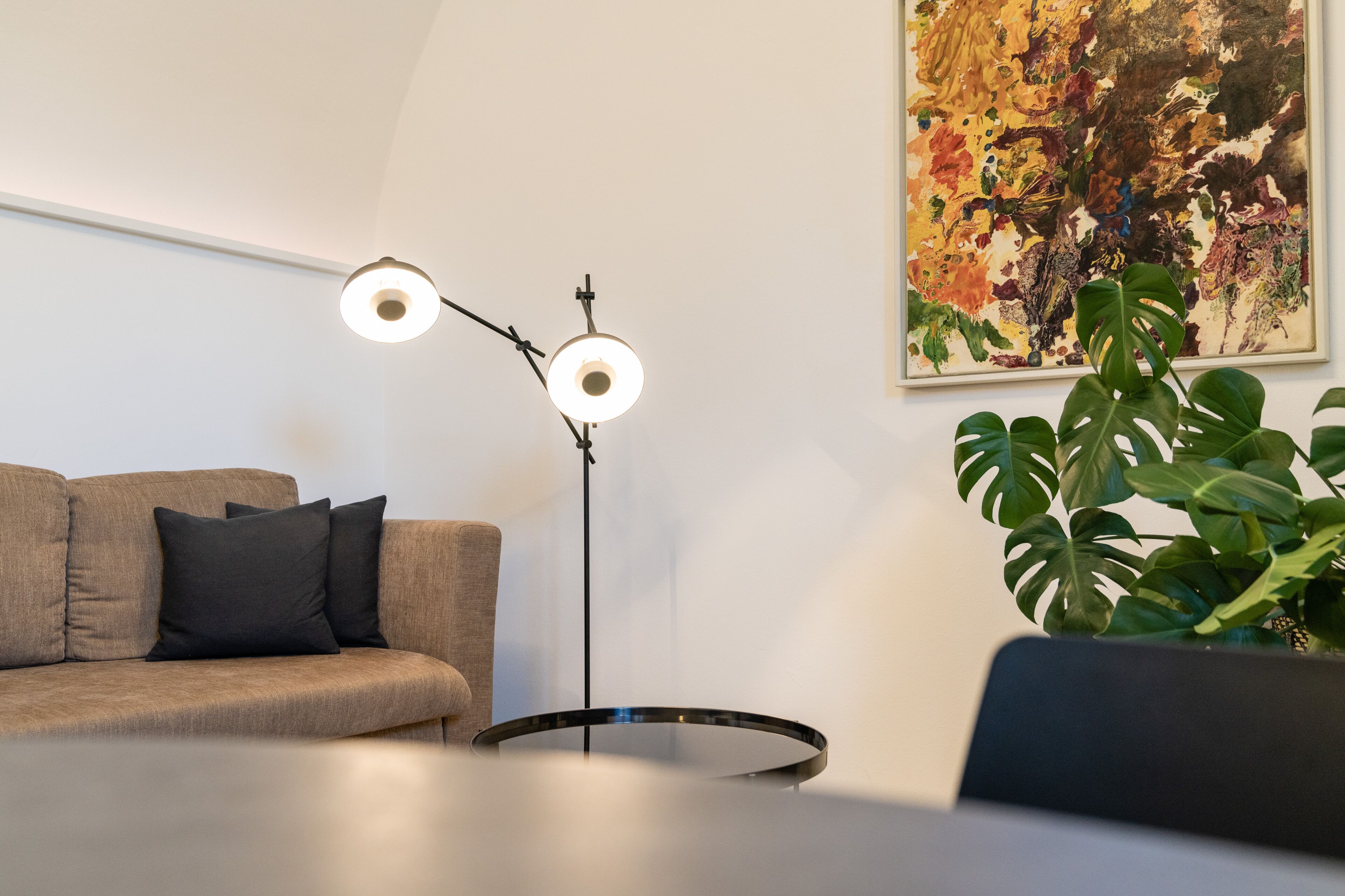 Uma sala de estar moderna com um abajur redondo, um sofá com almofadas, uma grande pintura abstrata e um vaso de planta.