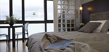 Quarto com uma cama grande, uma mesinha de cabeceira com um vaso de flores, uma cadeira e vista da cidade através de uma janela com painel de vidro decorativo.