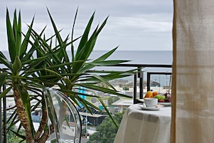 Uma varanda com uma planta em vaso, uma mesa com uma xícara e frutas, e vista para o mar.