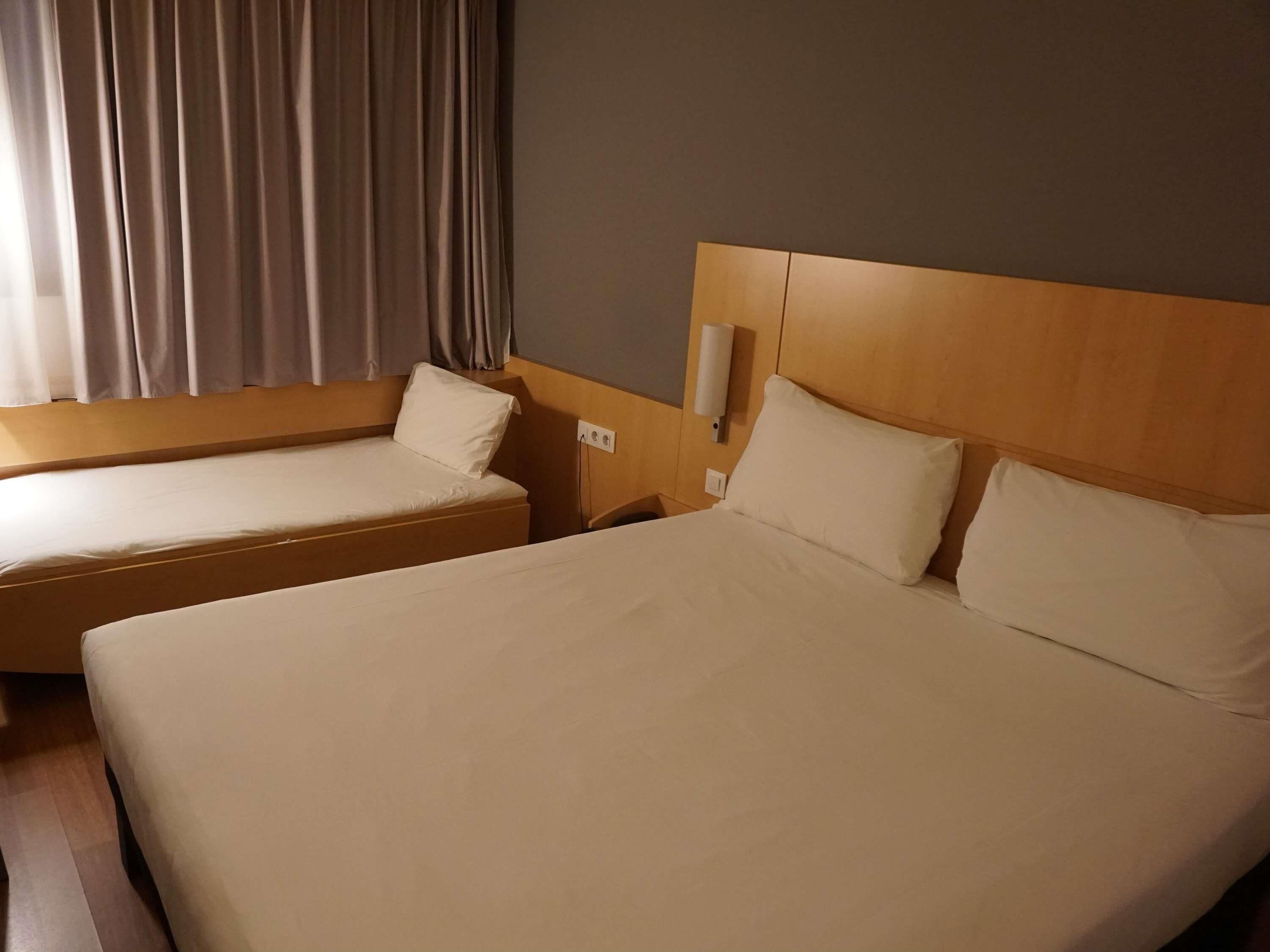 Quarto de hotel com duas camas de solteiro, cabeceira de madeira, luminária fixada na parede e janela com cortinas.