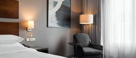 Quarto de hotel com uma cama grande, uma cadeira, uma escrivaninha, um abajur e um quadro na parede.