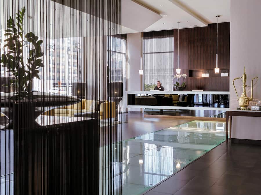 Um hall de hotel moderno com piso de vidro, balcão de recepção e uma grande janela com cortinas.