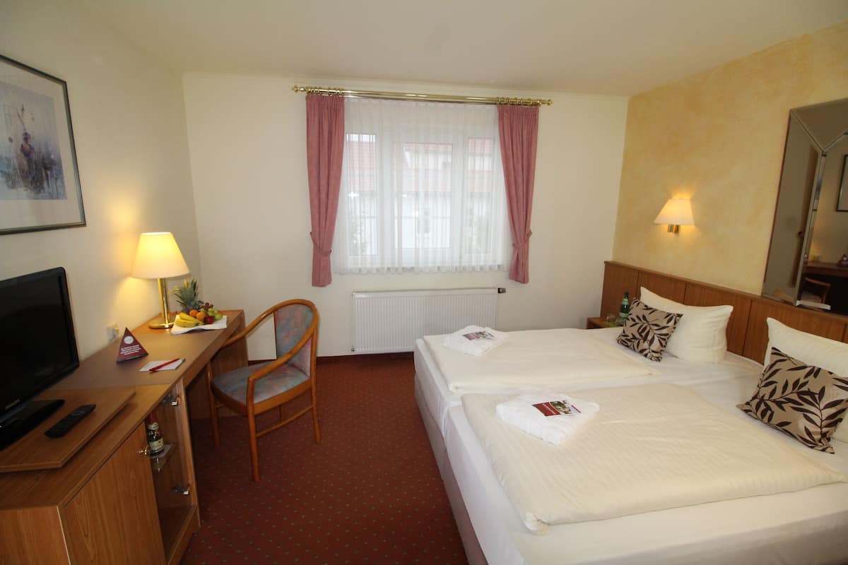 Quarto de hotel com duas camas, uma escrivaninha com abajur, uma cadeira, televisão e janela com cortinas.
