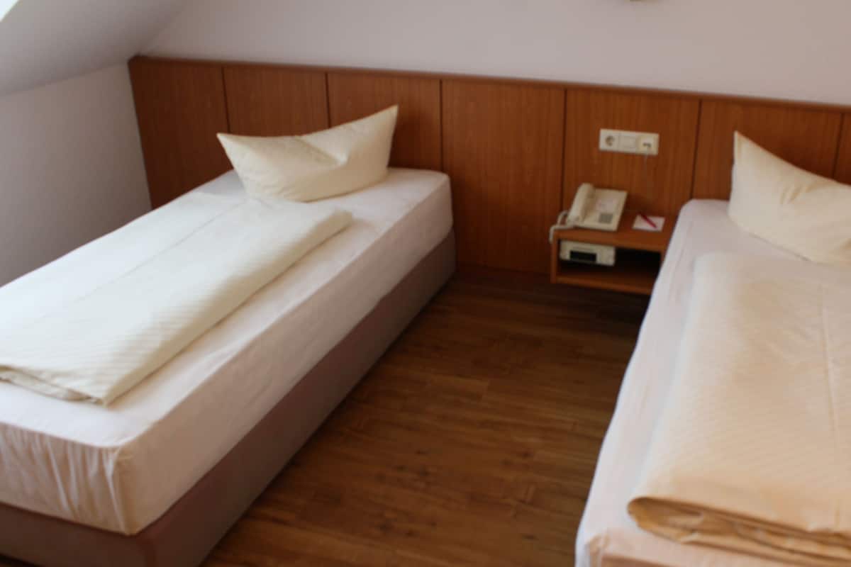 Quarto pequeno e compacto com duas camas de solteiro, cabeceiras de madeira, mesa de cabeceira com telefone e tomada na parede.