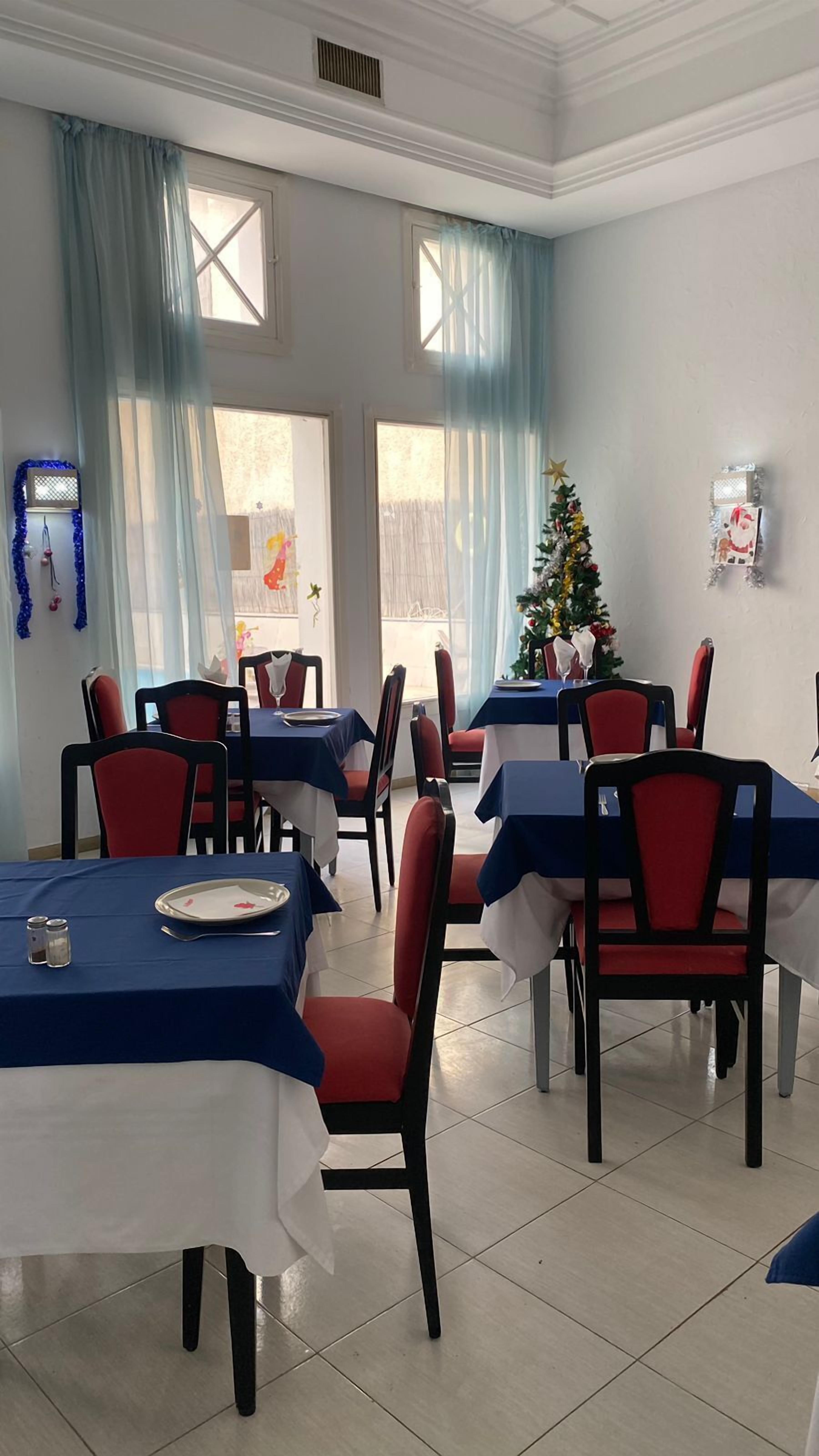 Uma área de refeições com mesas cobertas por toalhas azuis, cadeiras vermelhas e um pinheiro de Natal.
