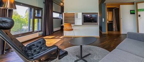 Quarto de hotel moderno com poltrona de couro, sofá cinzento, mesa redonda pequena e televisão.