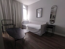 Quarto moderno e minimalista com sofá branco, uma mesa redonda pequena, uma cadeira de metal preta e um espelho sobre uma mesinha de cabeceira.