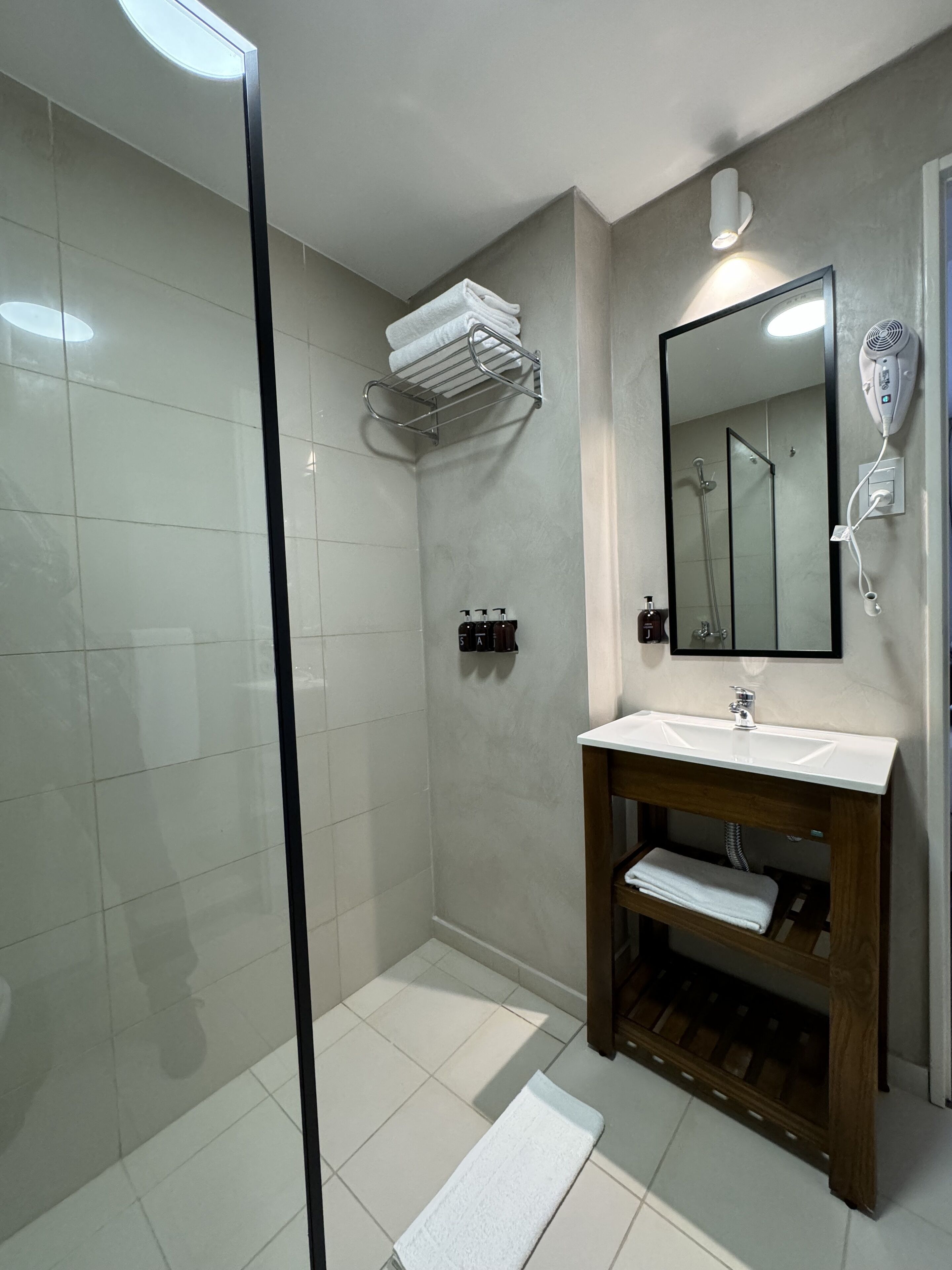 Um banheiro moderno com box de vidro, pia branca, toucador de madeira e espelho com secador de cabelo.