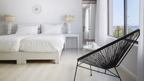 Quarto moderno com uma cama grande, luminárias de cabeceira, uma mesinha com uma cadeira e vista para o mar através de amplas janelas.
