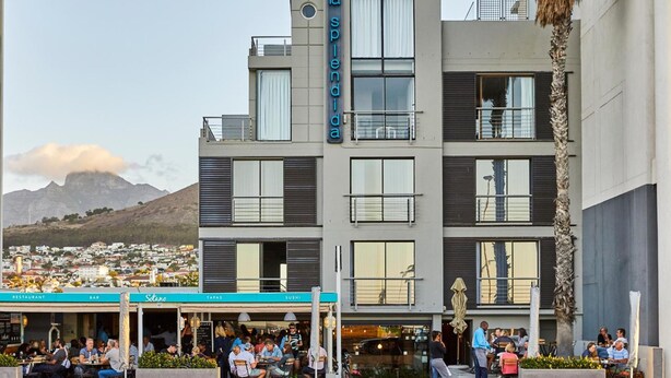 Um edifício de vários andares com um restaurante no andar térreo, com mesas ao ar livre e vista para uma paisagem montanhosa.