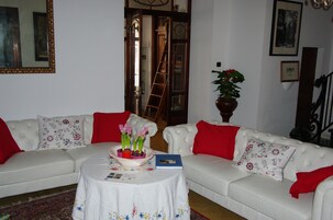 Uma sala de estar com dois sofás brancos, almofadas vermelhas, uma mesinha redonda com toalha florida e um vaso de planta.