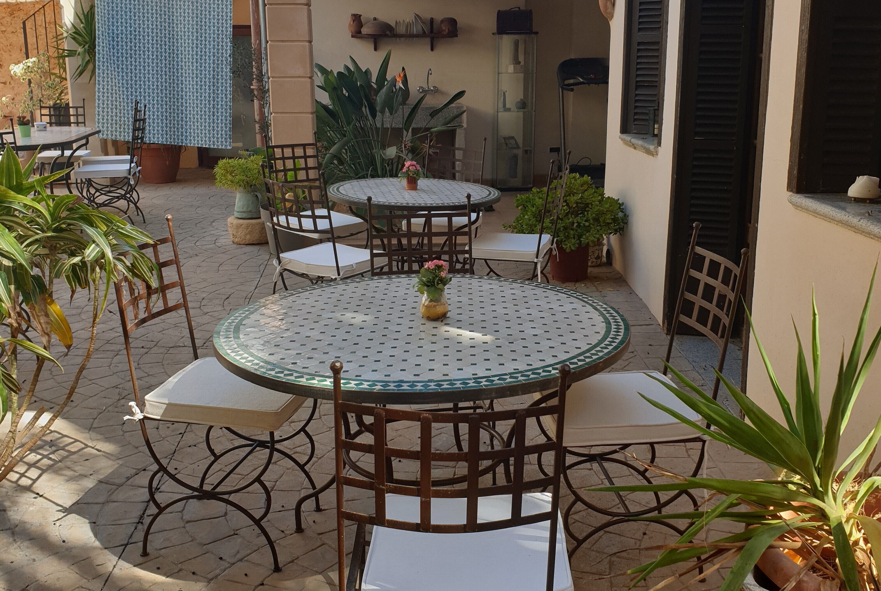 Um pátio externo com mesas e cadeiras redondas, cercado por plantas em vasos.