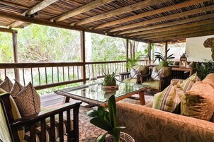Uma área de estar coberta ao ar livre, com mobiliário de madeira, mesas de vidro e plantas em vasos.