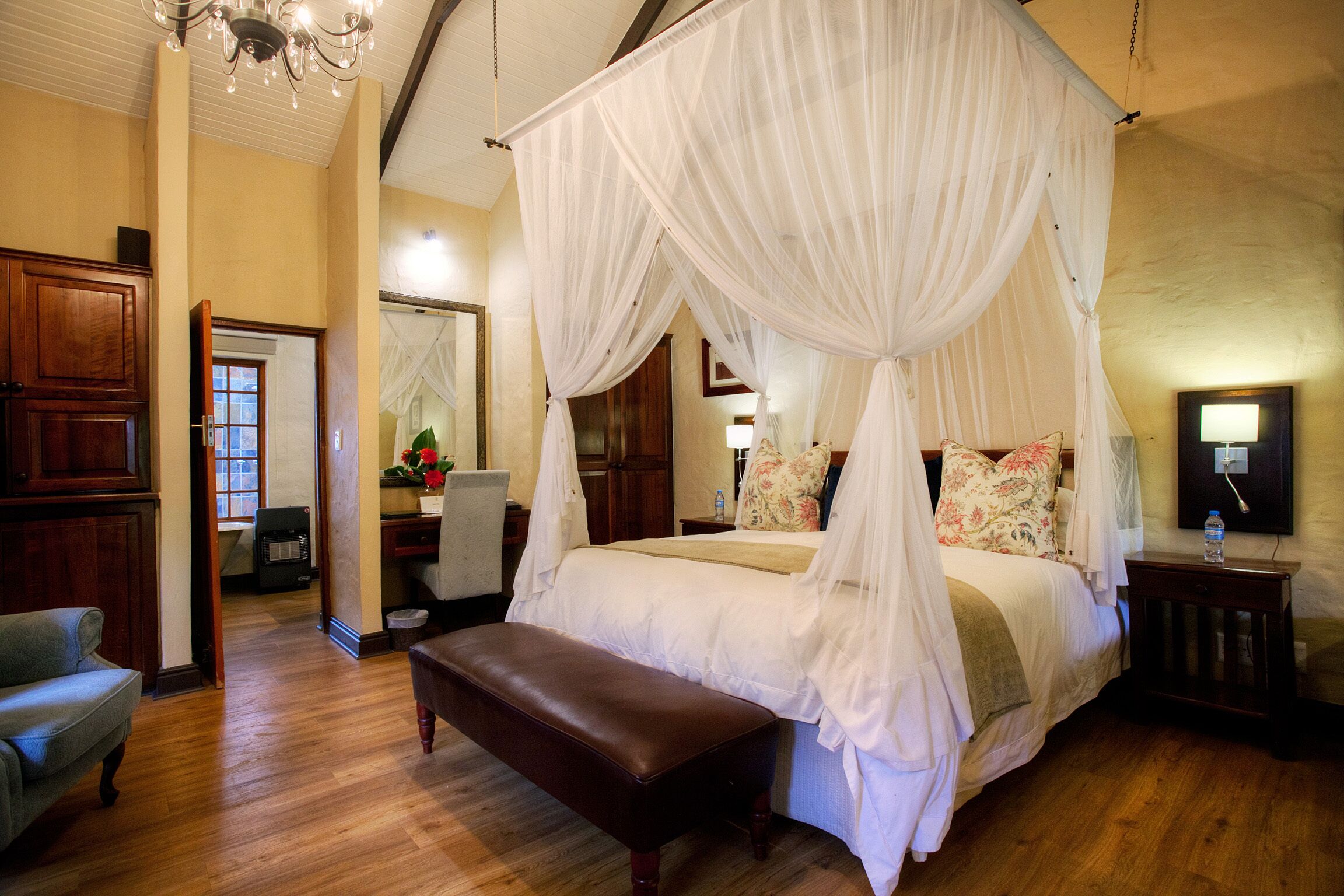 Quarto com cama de dossel, um banco, piso de madeira e um lustre.
