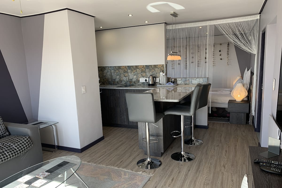 Quarto de hotel moderno com área de jantar, cozinha e espaço de estar com sofá e cadeiras.