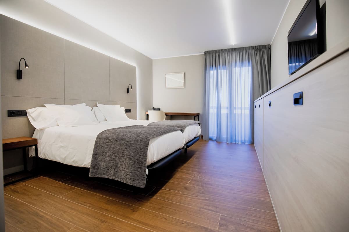 Quarto de hotel moderno com uma cama grande, mesas de cabeceira, televisão de tela plana e uma janela ampla com cortinas.