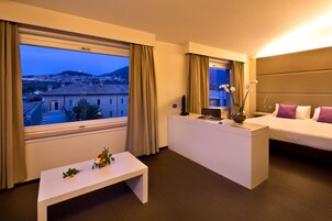 Quarto de hotel com uma cama, uma escrivaninha, uma mesinha com flores, um vaso e vista para a cidade através de amplas janelas.