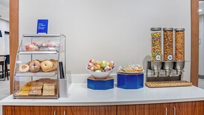 Um balcão de pequeno-almoço com diversos alimentos, incluindo pastelaria, uma taça de fruta e um dispensador de cereais.