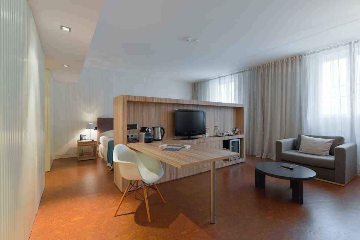 Quarto de hotel moderno com mesa de jantar de madeira, cadeiras brancas, sofá, TV de tela plana e máquina de café.