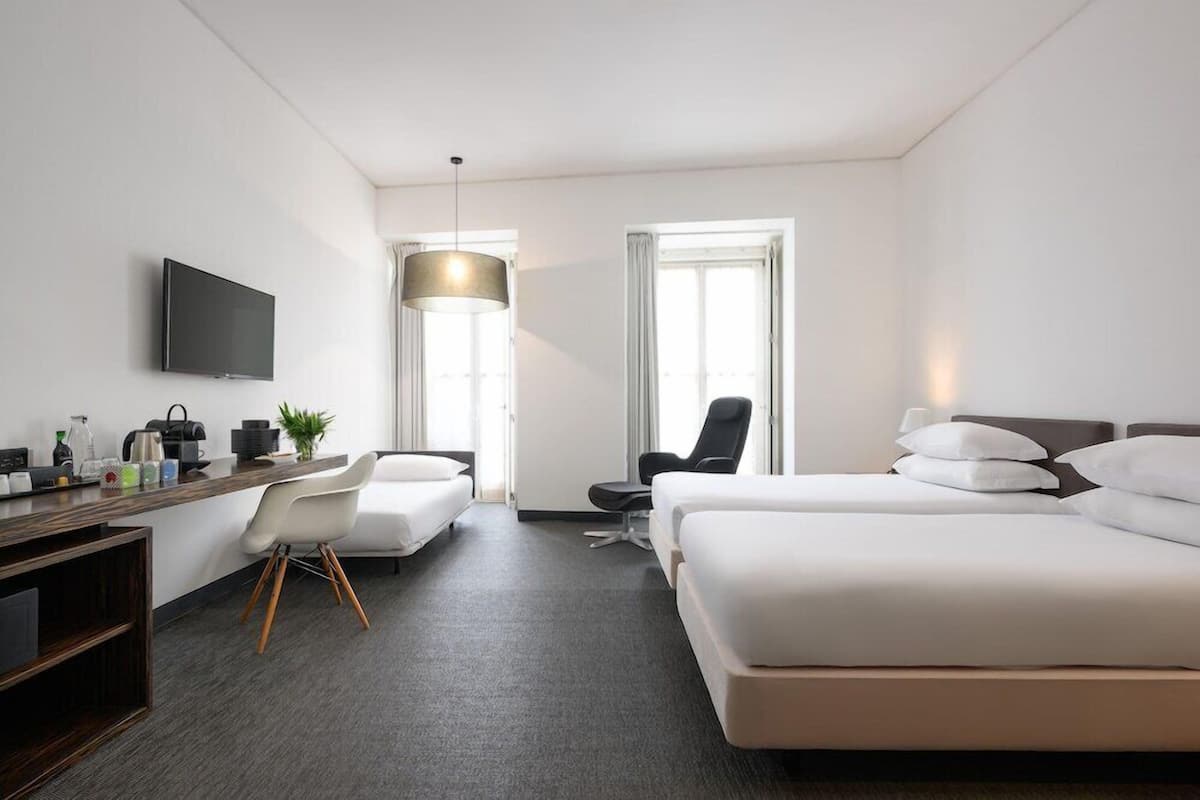Quarto de hotel com uma cama, uma escrivaninha, uma cadeira, uma televisão e uma janela.