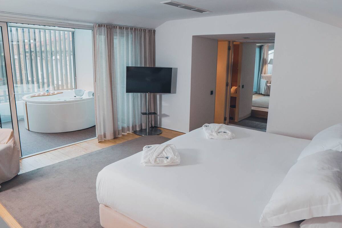 Quarto de hotel com uma cama grande, televisão de tela plana, banheira e vista para o exterior através de uma porta de correr de vidro.