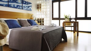 Quarto com uma cama grande, cabeceira de madeira, janela com vista e uma pequena mesa com uma planta e copos de vinho.