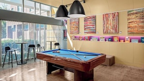 Uma mesa de bilhar em um espaço com janelas amplas, banquetas de bar e obras de arte coloridas nas paredes.