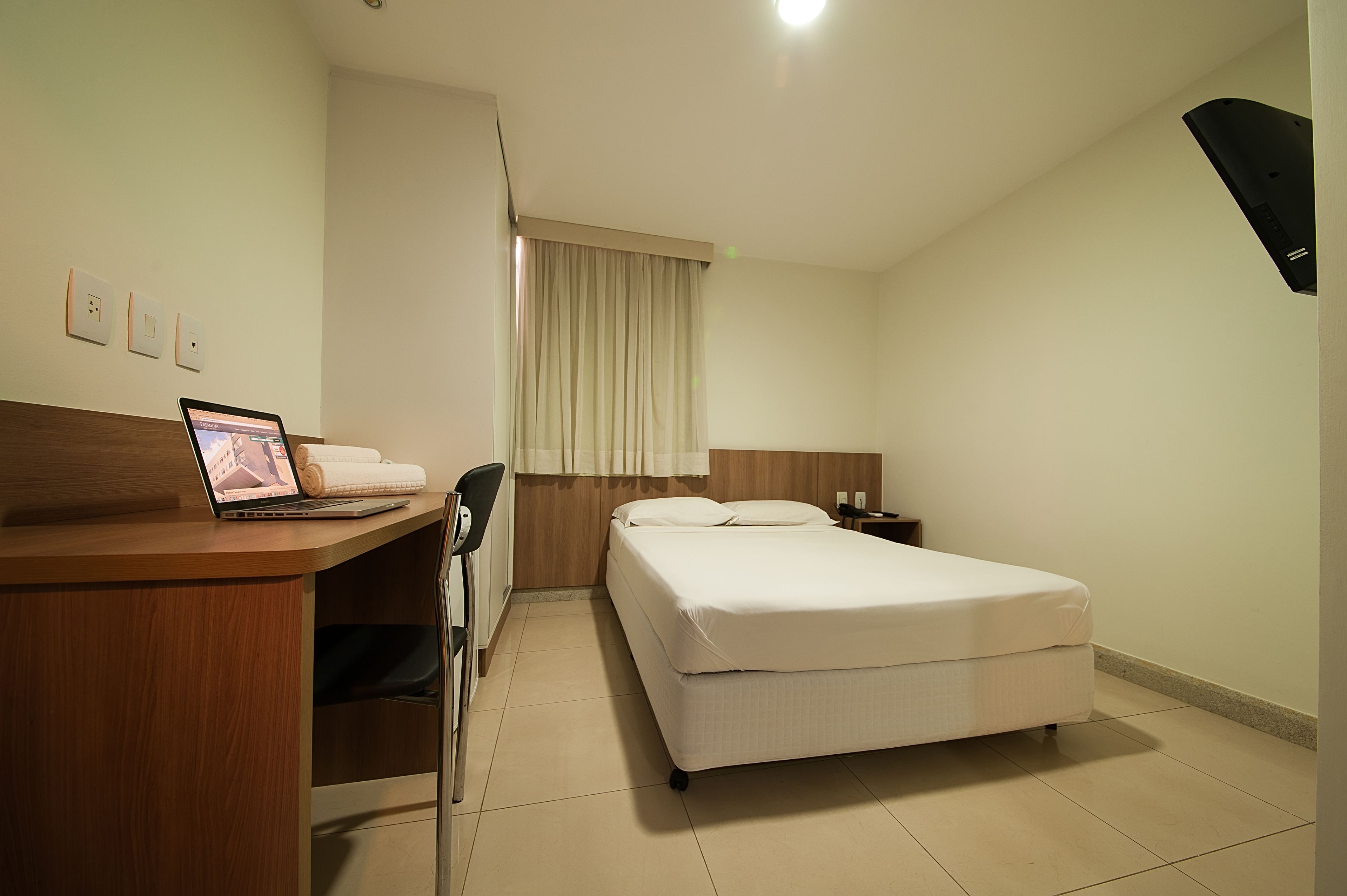 Quarto de hotel com cama, escrivaninha, laptop, cadeira, mesa de cabeceira e janela com cortinas.