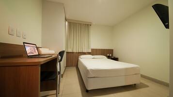 Quarto de hotel com cama, escrivaninha, laptop, cadeira, mesa de cabeceira e janela com cortinas.