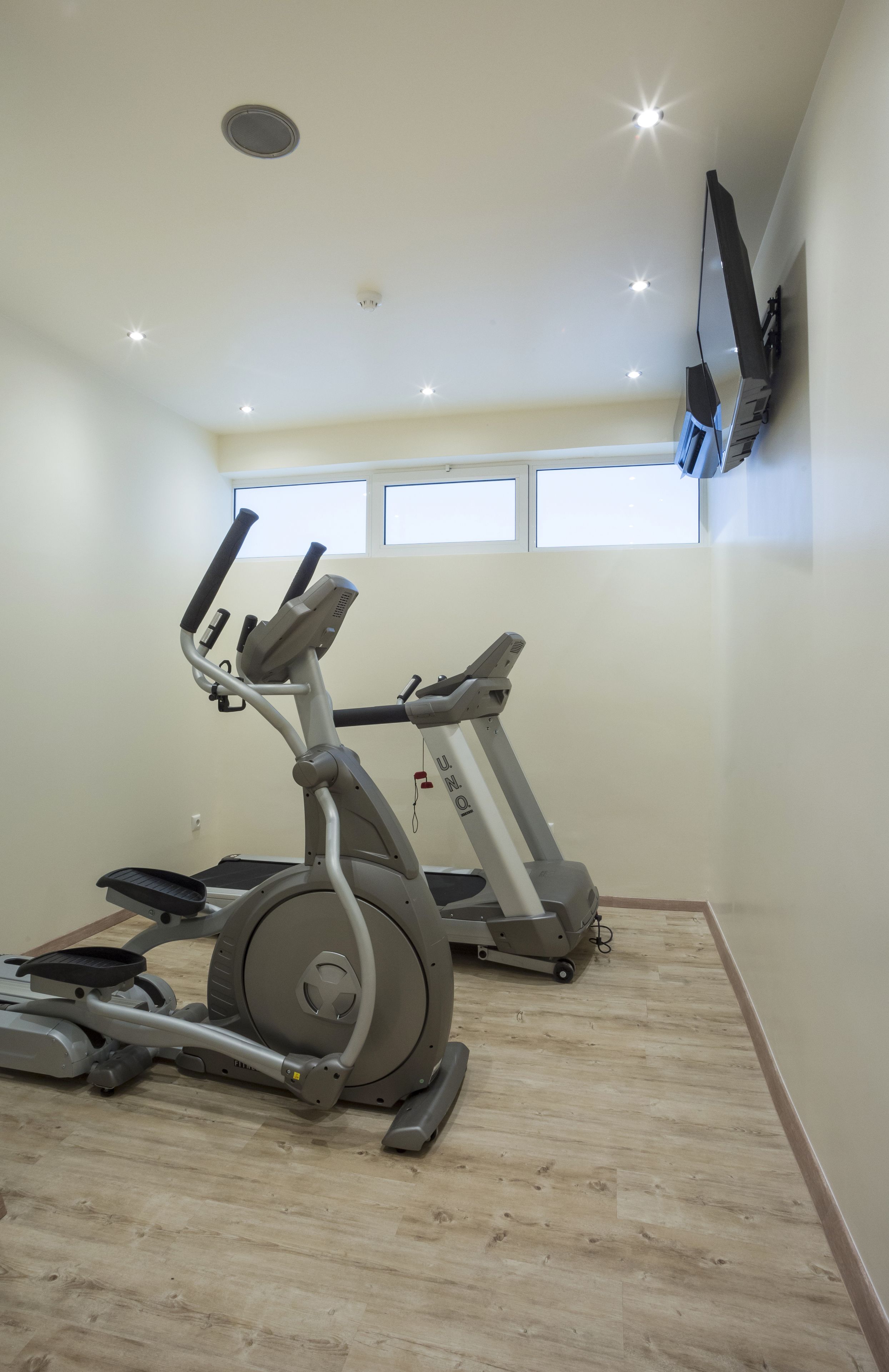 Uma academia em casa com duas bicicletas ergométricas e uma esteira, uma TV fixada na parede, e uma janela que traz luz natural.